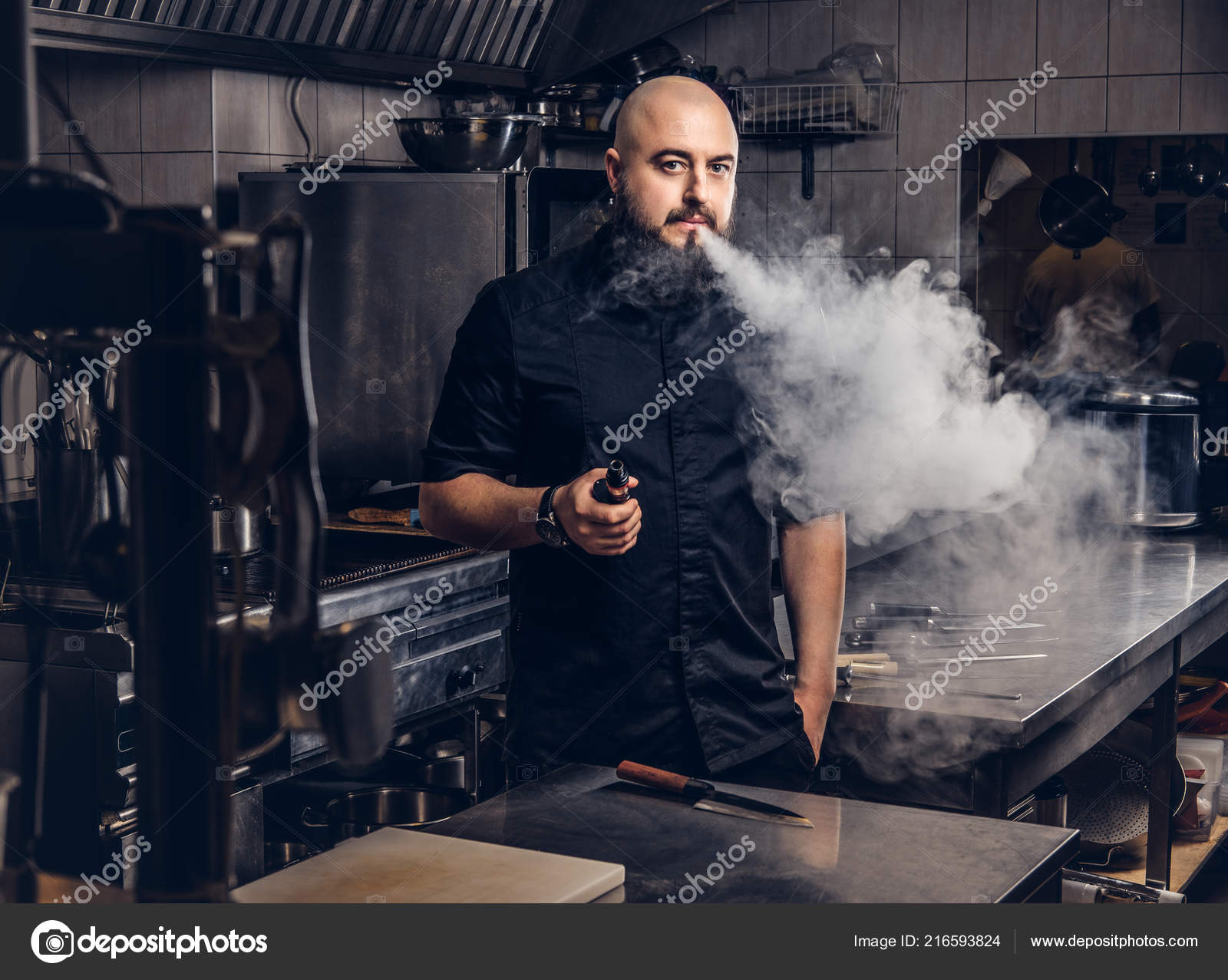 Bearded chef in black uniform smoking e-cigarette while standing in the ...