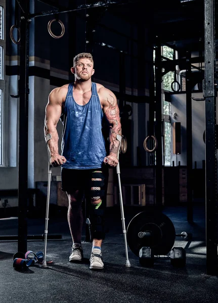 Muscular bodybuilder standing with crutches and bandage on a leg in a ...