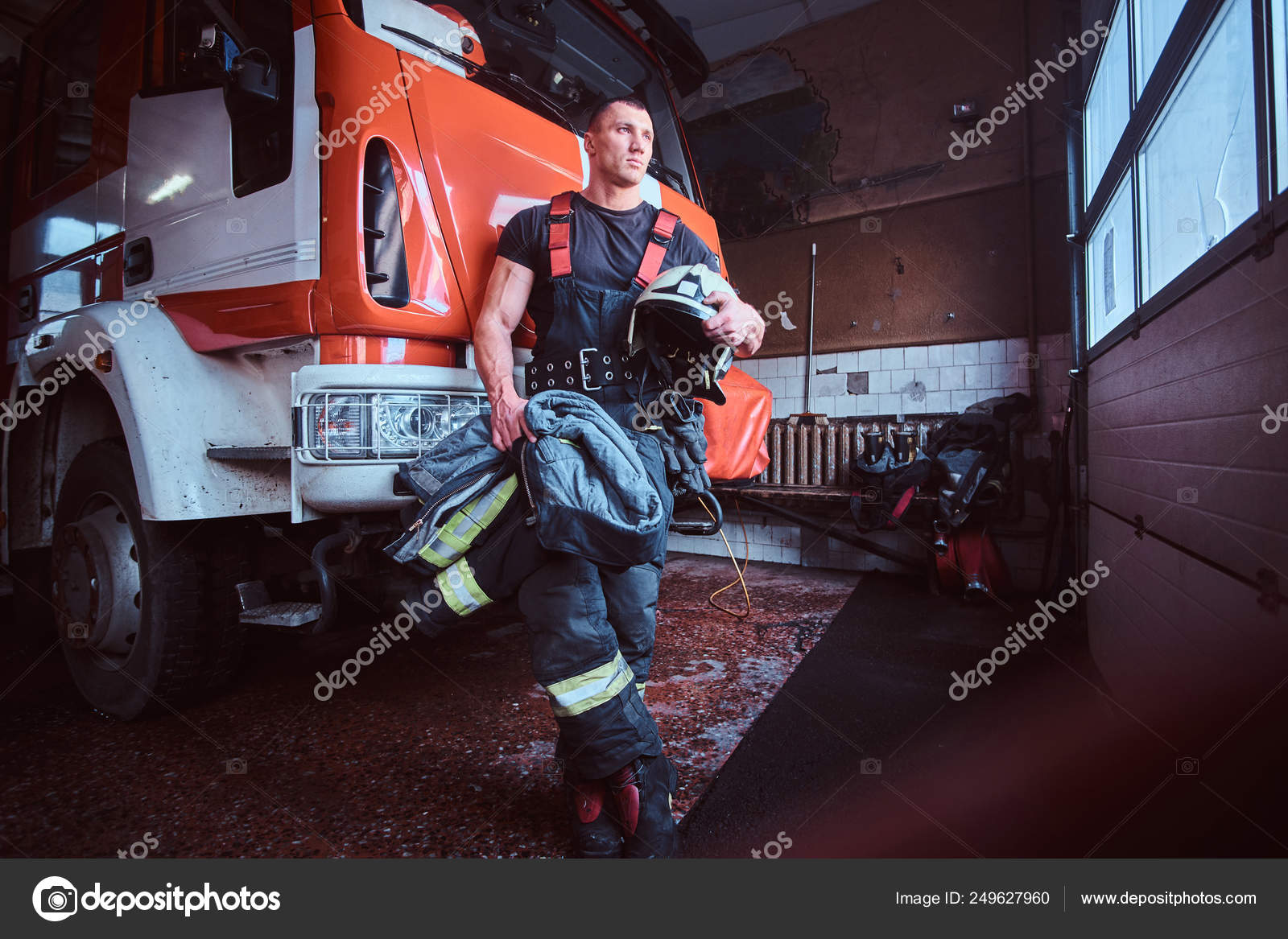 Young fireman holding a jacket and protective helmet in a garage of a ...