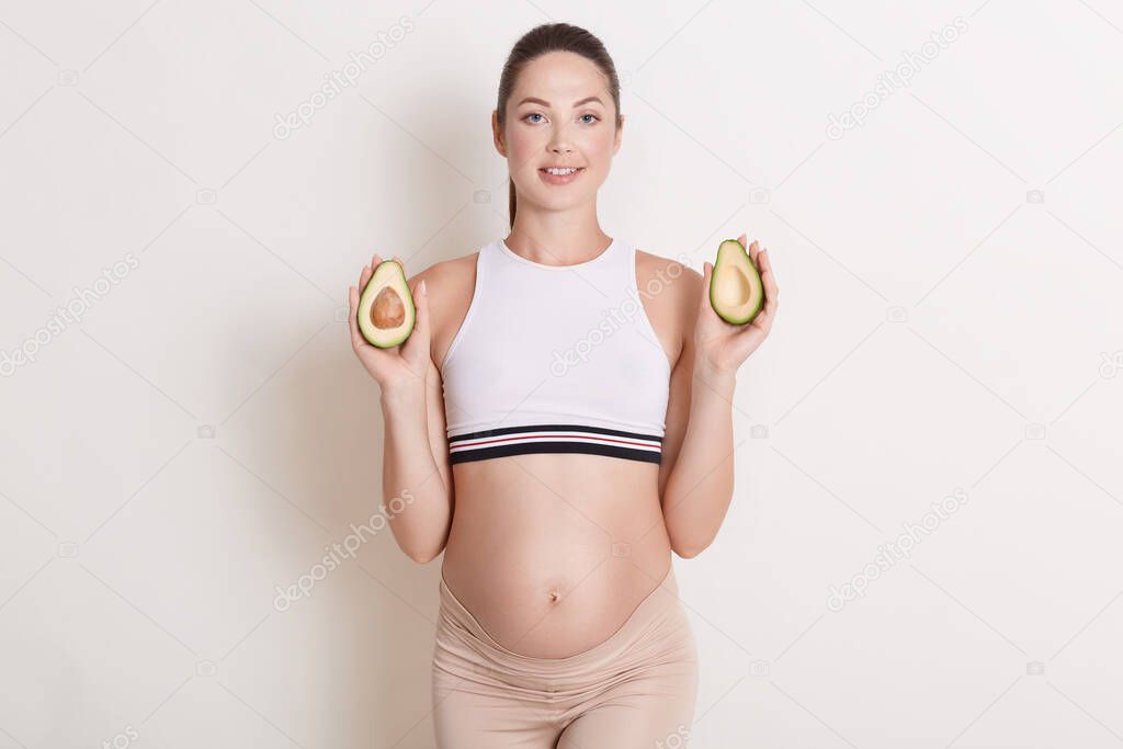 Mujer embarazada joven sosteniendo dos mitades de aguacate fresco mientras posa contra la pared ...