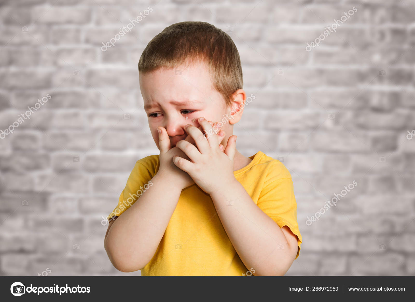 Crying baby boy in a yellow T shirt covers his face with hands and ...