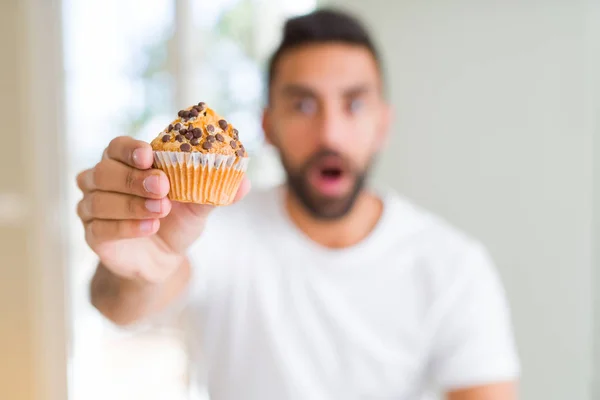 Handsome hispanic man eating chocolate chips muffin scared in shock ...