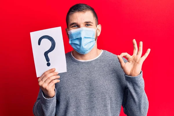 Young handsome man wearing medical mask holding question mark doing ok ...