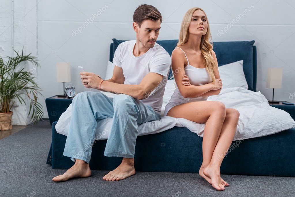 joven con tel fono inteligente mirando novia molesta sentado con los brazos cruzados en la cama ...