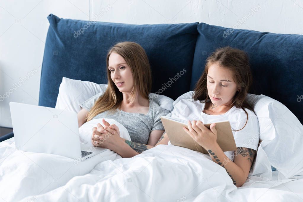 Dos Lesbianas Con Portátil Y Libro Tomados De La Mano Mientras Yacen