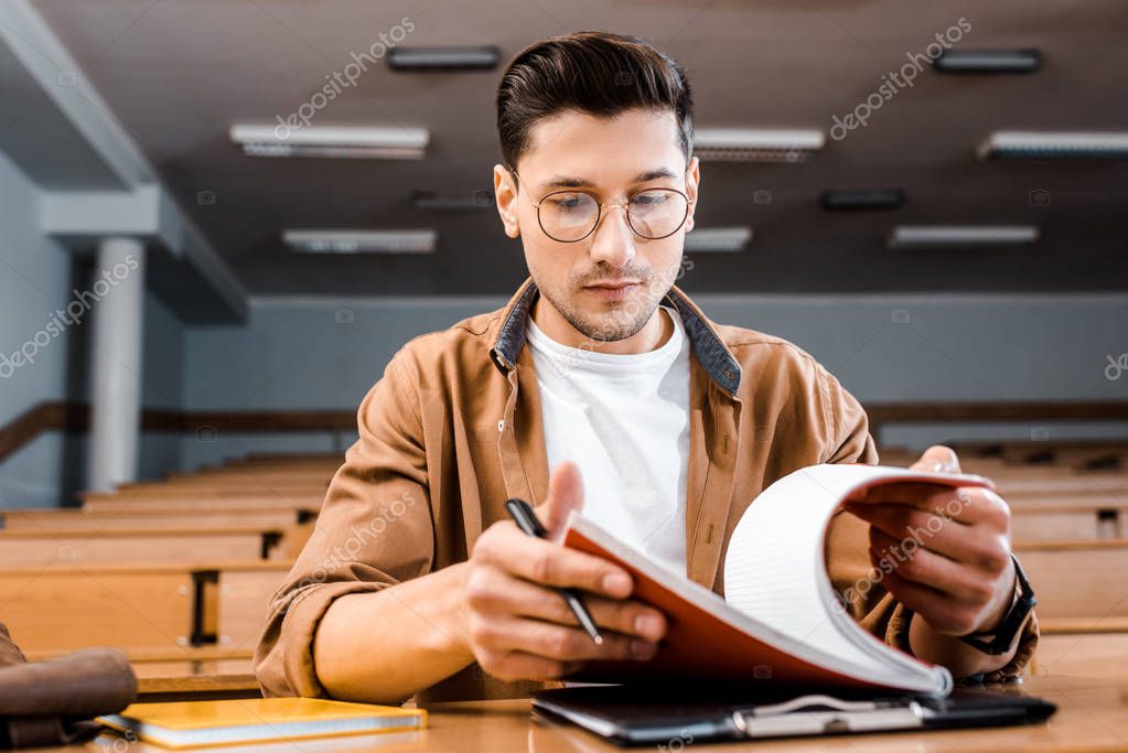 estudiante masculino concentrado en gafas sentado en el escritorio y sosteniendo un cuaderno con ...