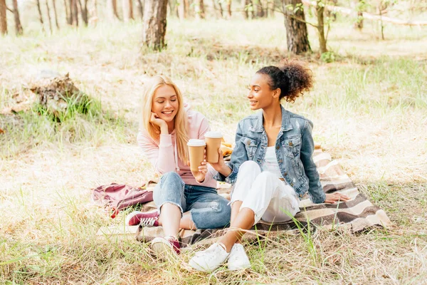 two multiethnic friends sitting on plaid blanket and holding paper cups ...