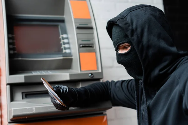 Side view of robber in balaclava holding credit cards near automated ...