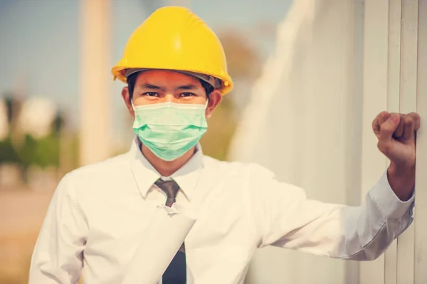 Asian man Engineer use face mask yellow hard hat holding blueprint ...