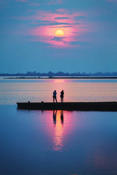A romantic sunset with people on a jetty at the lake together watching ...