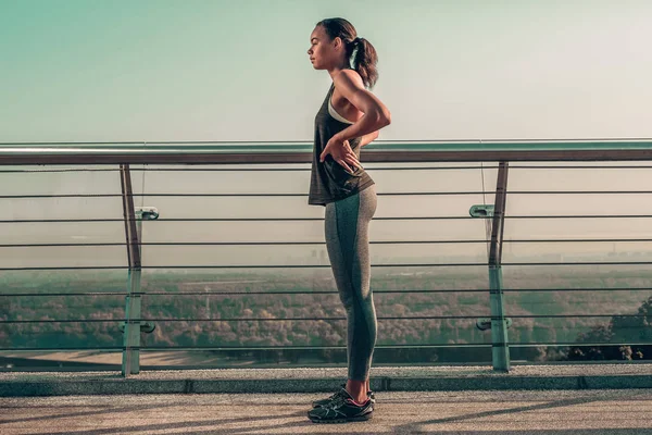 Calm young athlete in sports clothes resting on the bridge and looking ...