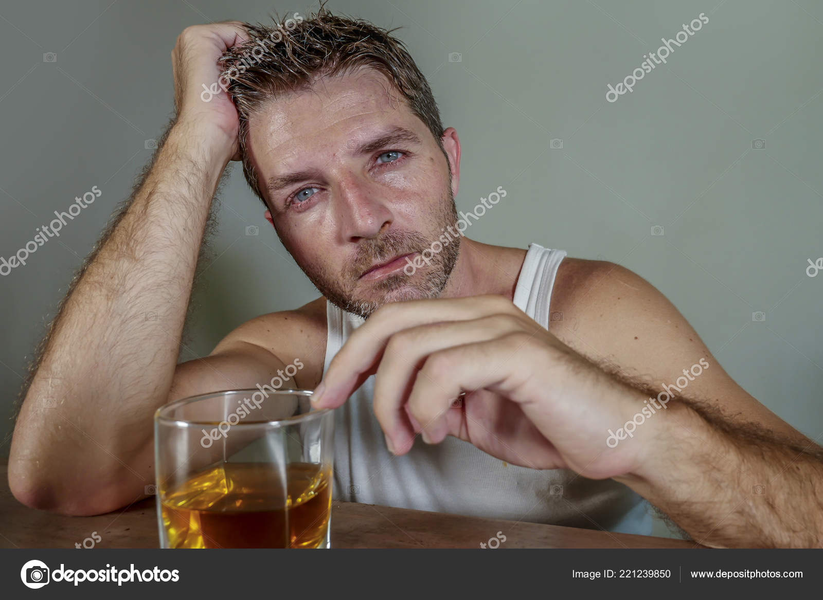 Young wasted and depressed alcohol addict man in dirty singlet drinking glass of whiskey feeling ...