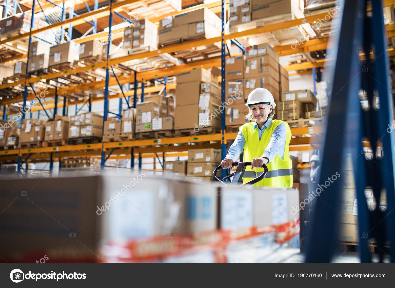 A senior woman warehouse worker pulling a pallet truck with boxes ...