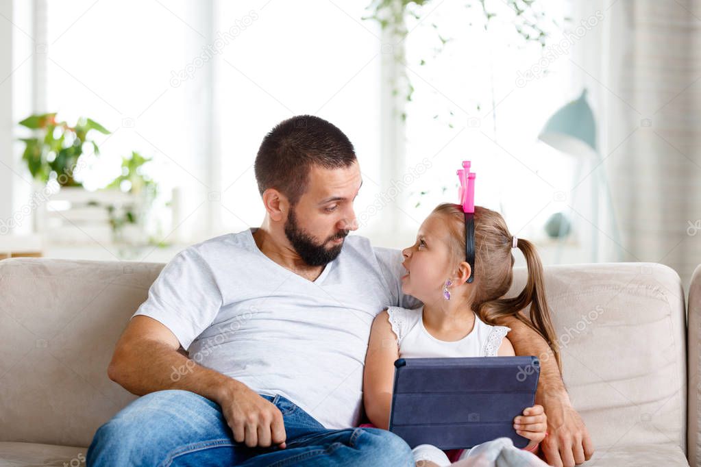 Padre e hija sentados en el sofá en casa, usando tableta digital juntos y haciendo caras ...