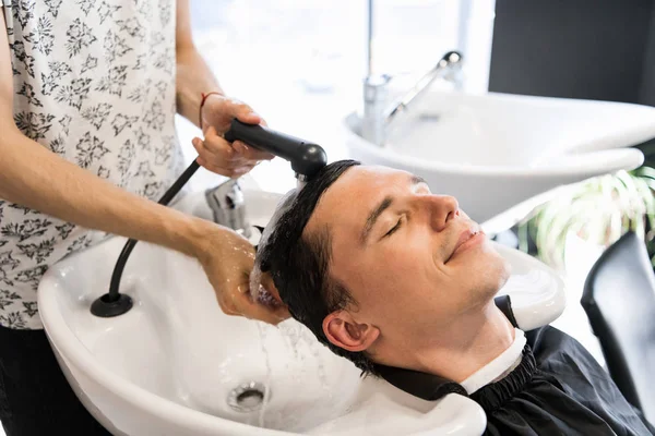 Relaxed young man with eyes closed leaning on sink while hairdresser washing his hair in salon Stock Image Everypixel