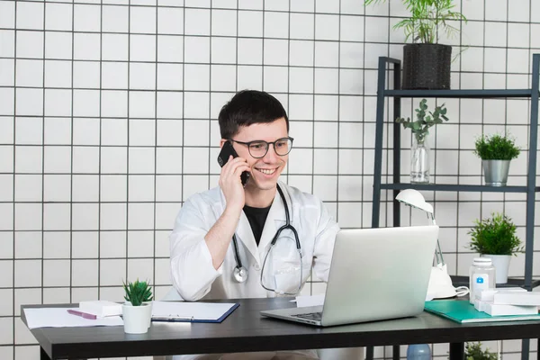 Male doctor using telephone while working on computer at table in ...