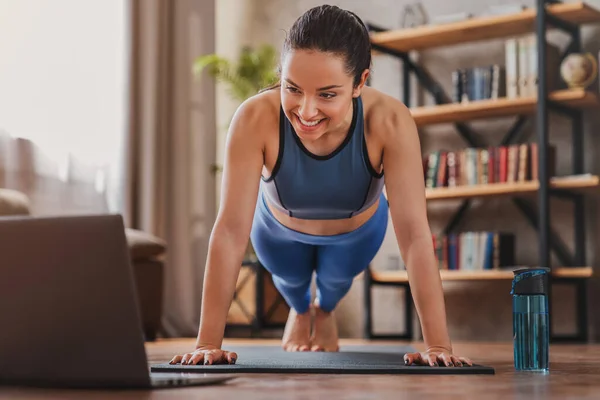 Smiling young woman watching training videos on laptop while exercising ...