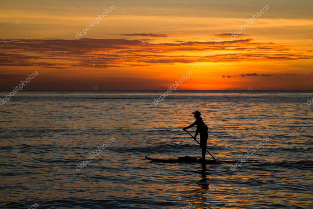 Silueta del joven remando en un tablero de SUP en el mar al atardecer ...