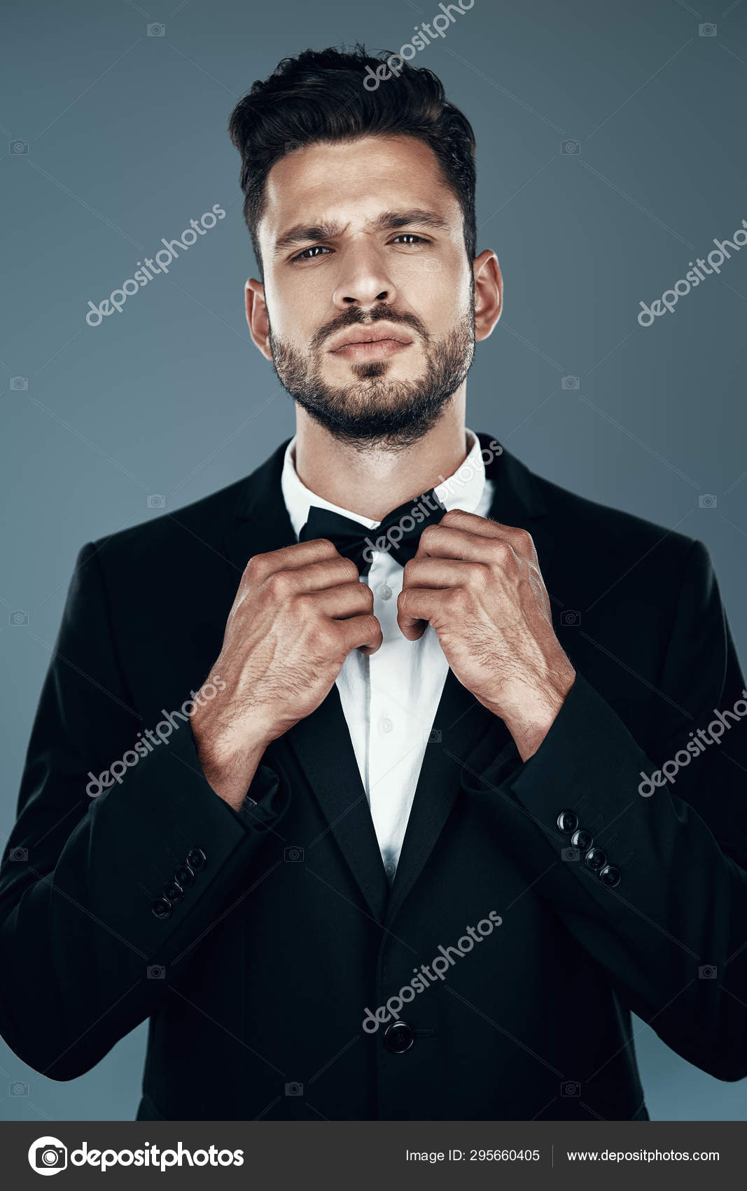 Charming young man in full suit looking at camera and adjusting bow tie ...