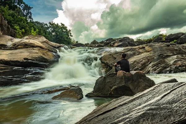 hombre sentado en la roca observando el hermoso arroyo de cascada en la ...
