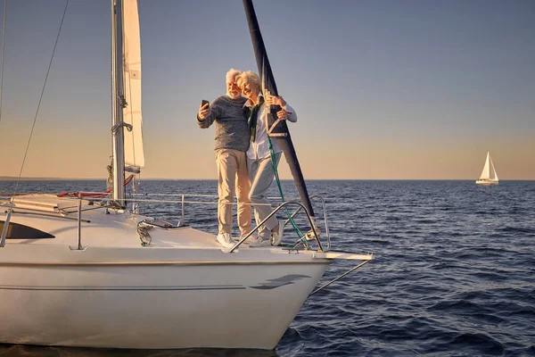 Full length of a happy senior couple standing together on the side of sailboat floating in sea ...