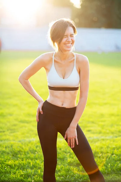 Laughing fitness woman with perfect figure wears fashionable sportswear posing near the grass ...