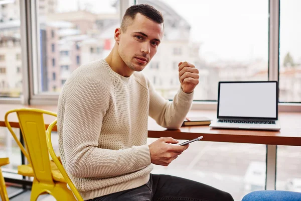 Portrait of handsome young man holding smartphone and updating profile in social networks on ...