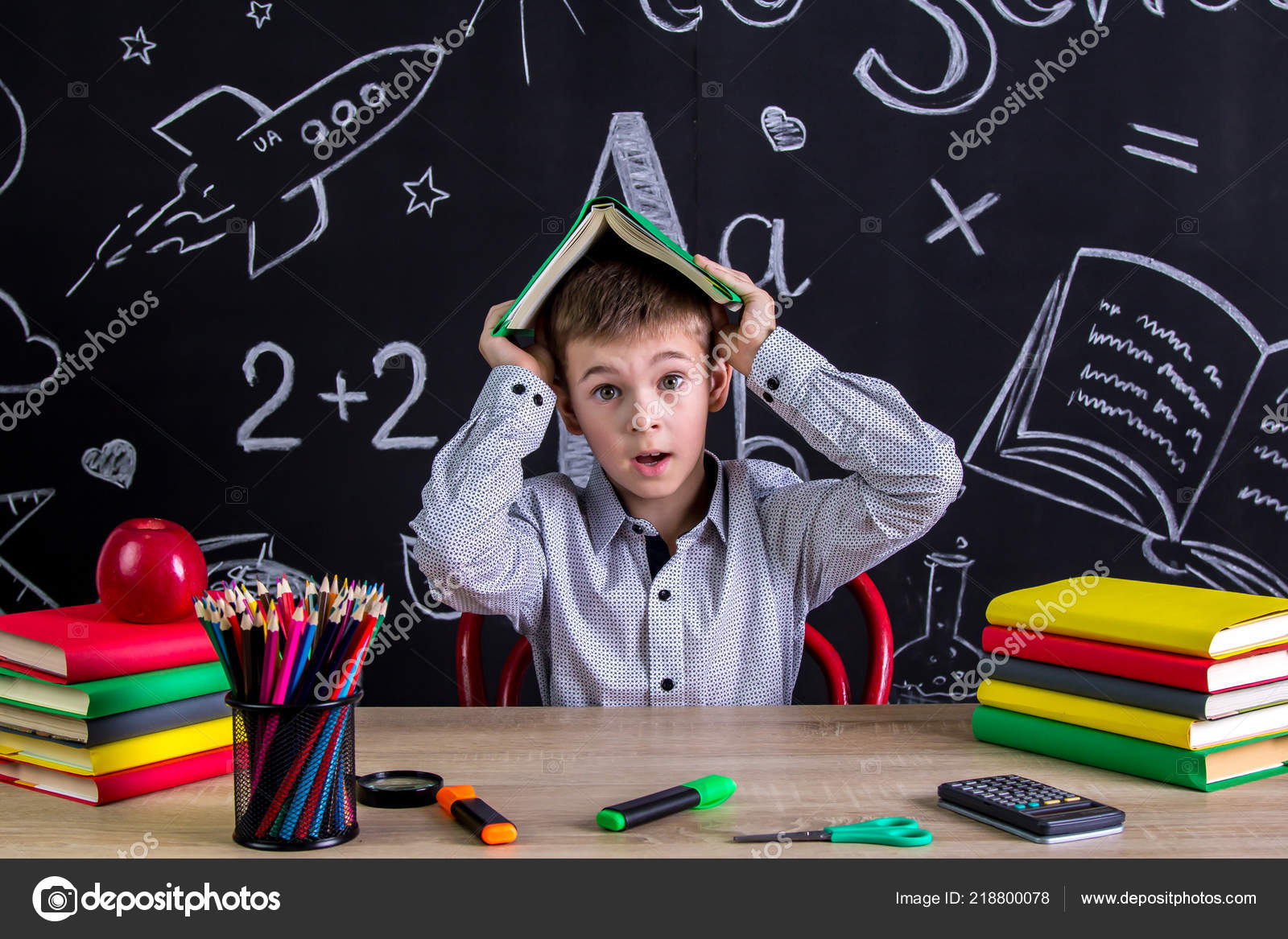 Scared and puzzled schoolboy sitting at the desk holding the book above ...