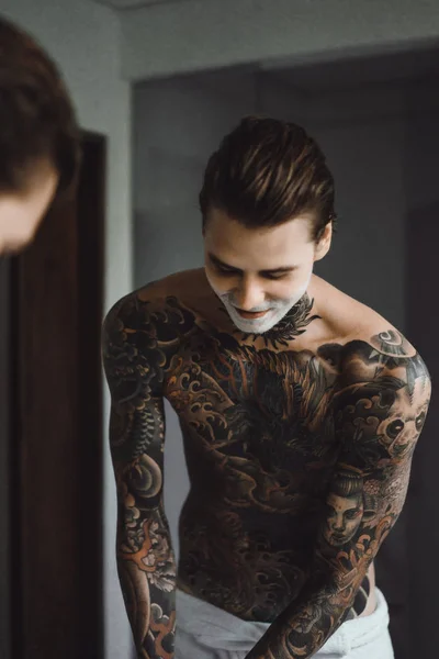 a young handsome tattooed man in the morning shaves in front of a ...