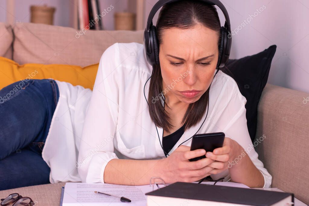 Bonita joven morena aburrida con su smartphone estudiando estirada en el sof en casa escuchando ...