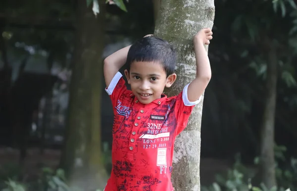 Lindo niño parado al lado de un árbol afuera en un cálido día soleado y posando para la ...