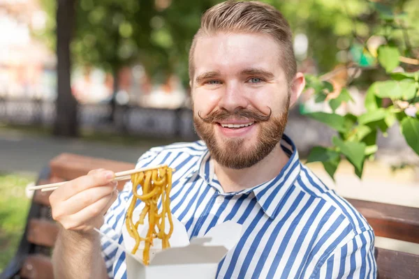 Young male hipster with a mustache and beard is eating chinese noodles ...