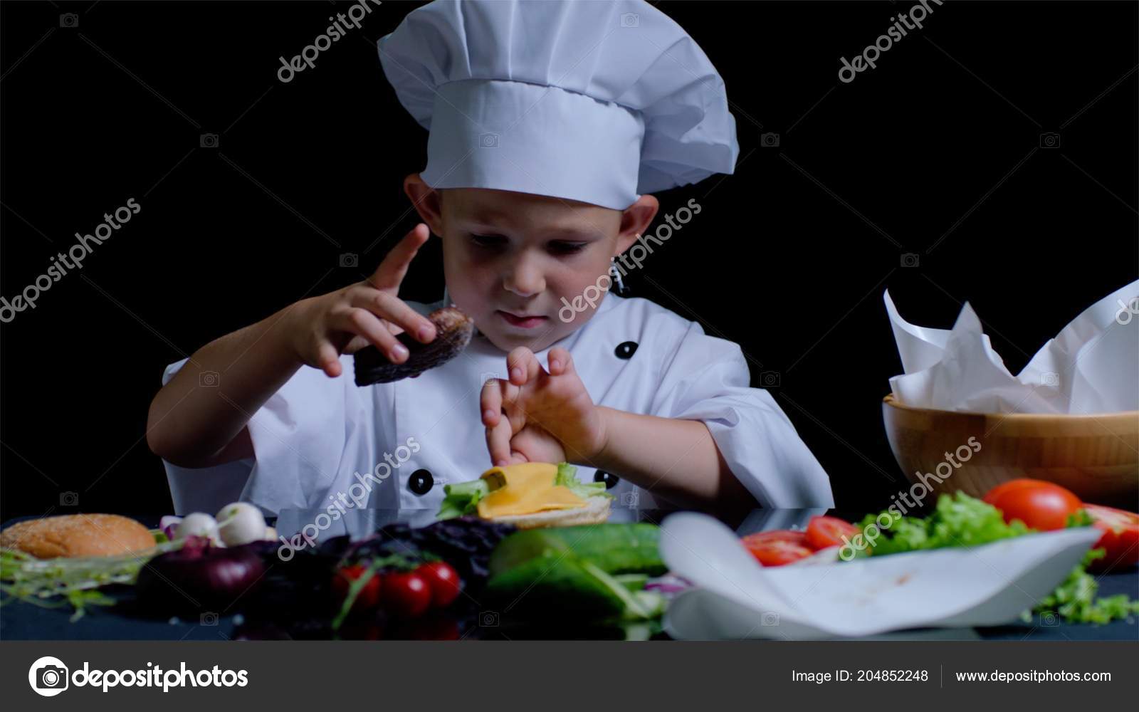 Little boy is cooking a burger at the kitchen, he is wearing chefs suit ...