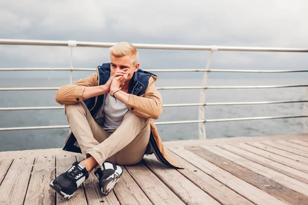 Upset young man sitting by sea on pier in Odessa thinking on rainy ...