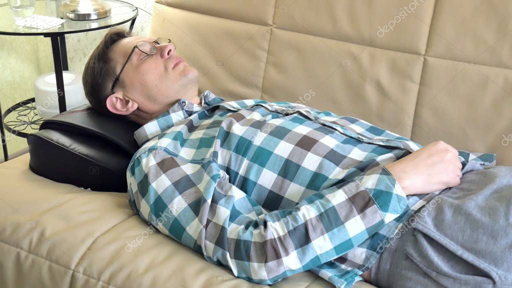 hombre se encuentra en el sofá en casa, bajo la cabeza de un masajeador ...